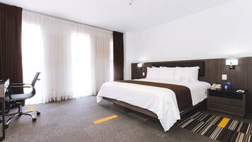 Quarto de hotel com uma cama grande, uma escrivaninha com cadeira, uma mesa de cabeceira com telefone e uma janela com cortinas.