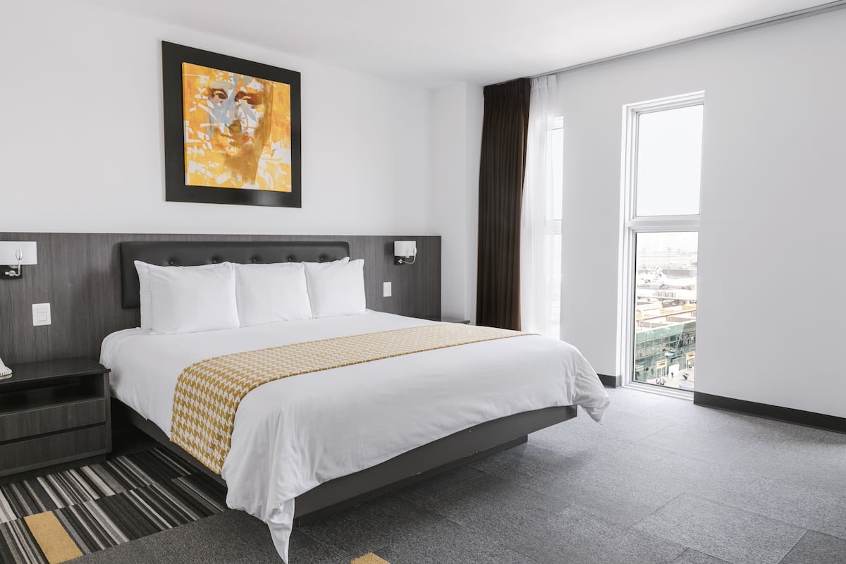 Quarto de hotel com uma cama grande, uma mesinha de cabeceira, uma luminária na parede, uma obra de arte emoldurada e uma janela com vista para prédios.