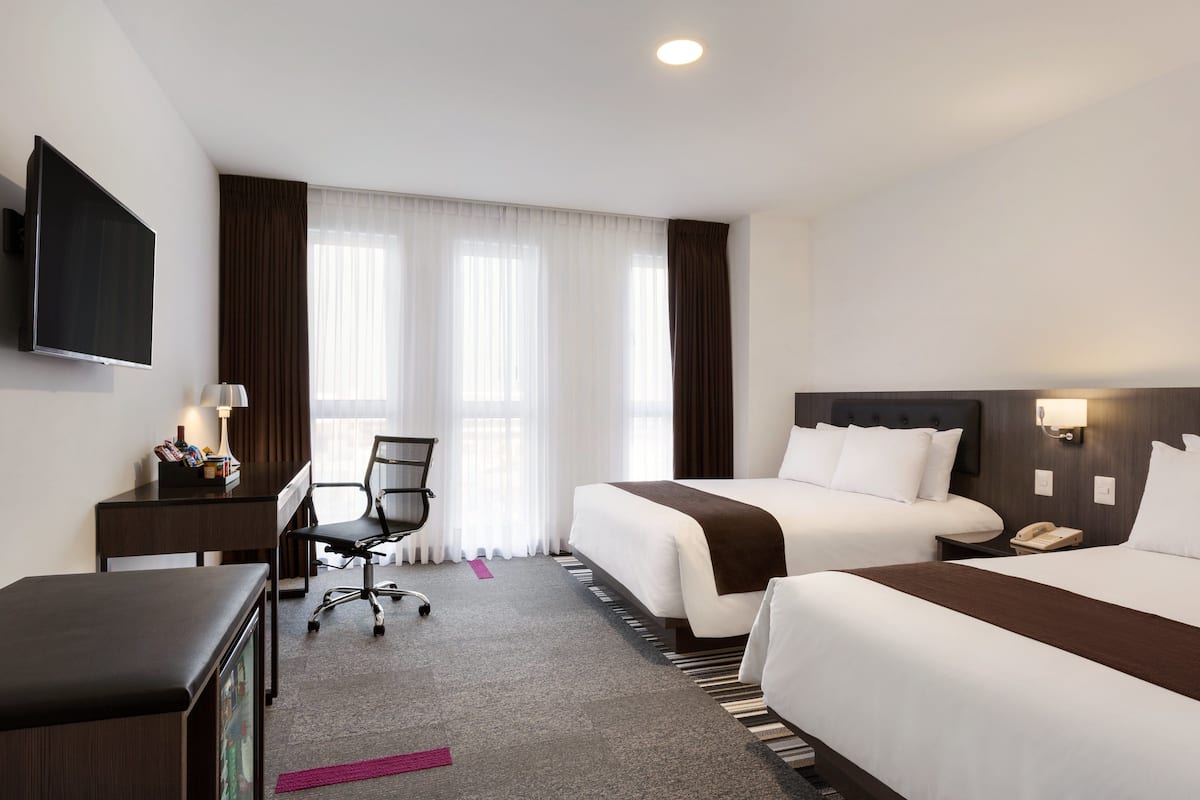 Quarto de hotel com duas camas, uma escrivaninha, uma cadeira, uma televisão e uma janela com cortinas.