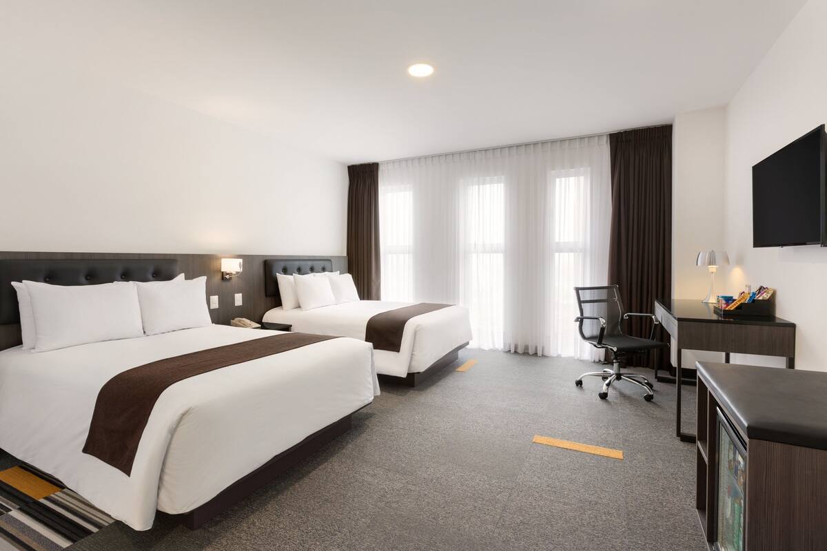 Quarto de hotel com duas camas, uma escrivaninha, uma cadeira, uma televisão e uma janela com cortinas.