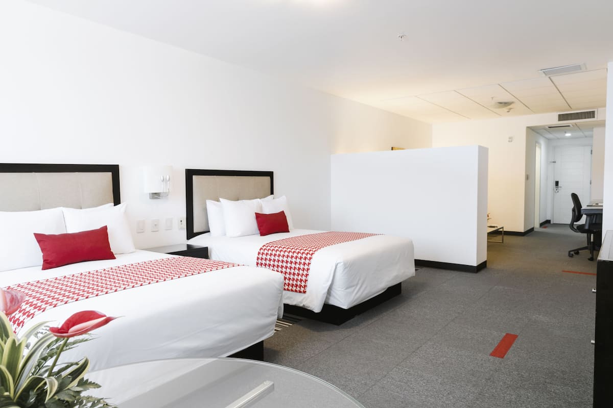 Quarto de hotel com duas camas, cada uma com roupa de cama branca e detalhes vermelhos, uma escrivaninha e uma cadeira.