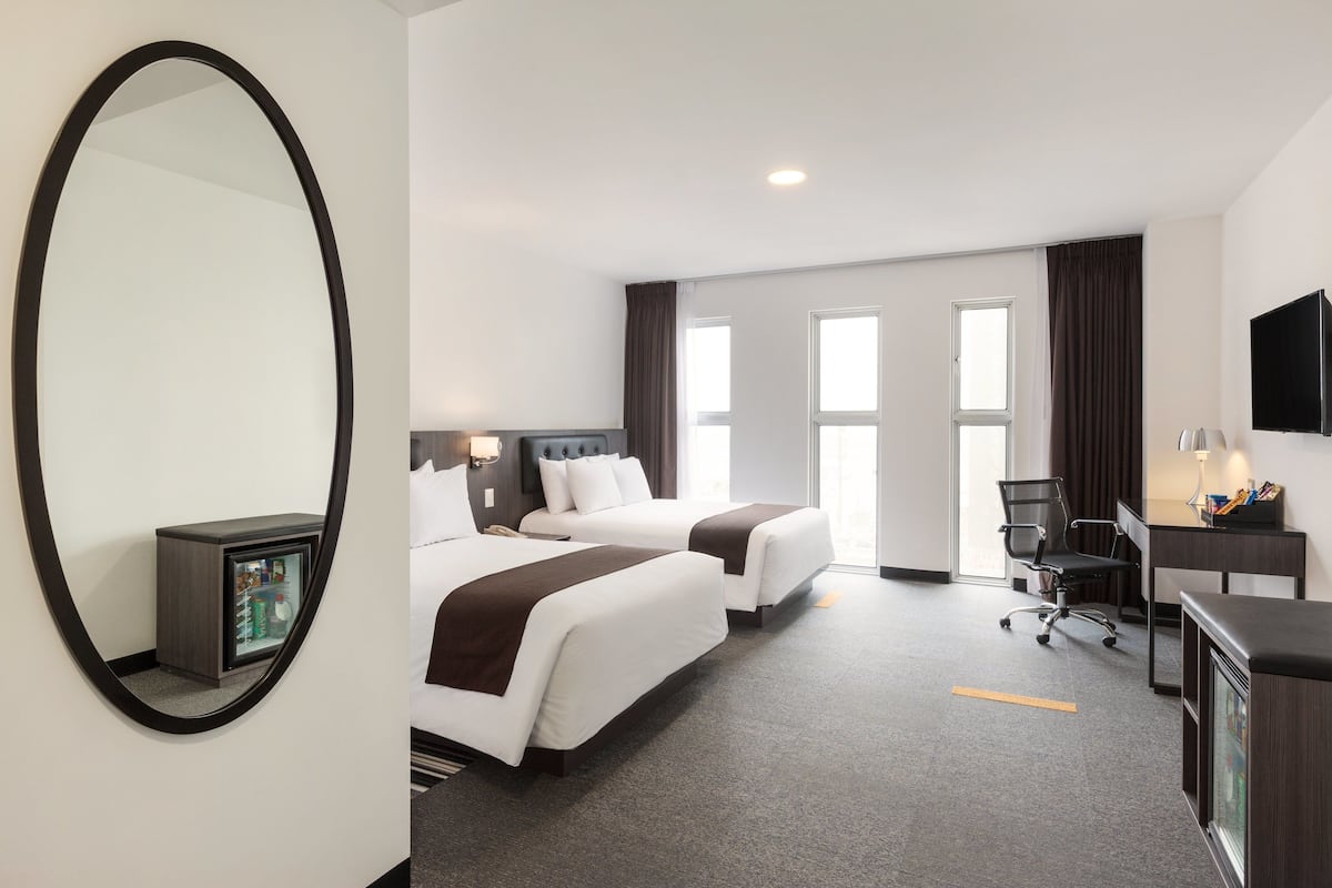 Quarto de hotel com duas camas, uma escrivaninha, uma cadeira, televisão e um espelho grande.