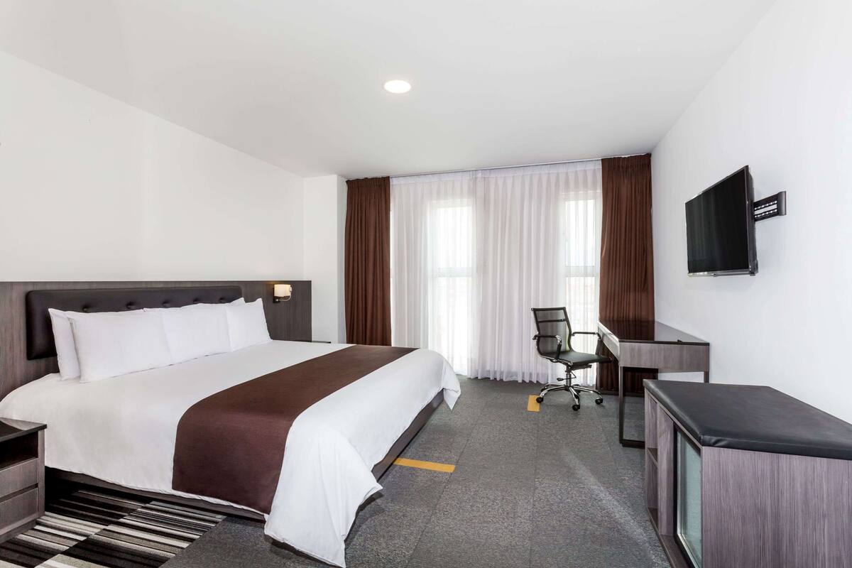 Quarto de hotel com uma cama grande, uma escrivaninha, uma cadeira, uma televisão e uma janela com cortinas.