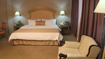 Quarto de hotel com uma cama grande, duas luminárias de cabeceira, uma cadeira e uma mesinha com telefone e planta.