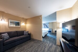 Quarto de hotel com sofá cinza escuro, televisão de tela plana, cama e uma janela grande.