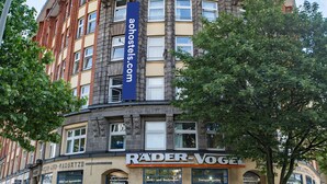 Um edifício de vários andares com uma placa chamativa da 'a&o', uma loja 'RADER VOGEL' e vários carros estacionados.
