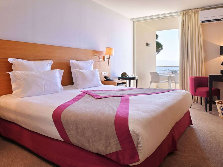Quarto de hotel com uma cama grande, uma escrivaninha com cadeira, uma mesinha e vista para o exterior.