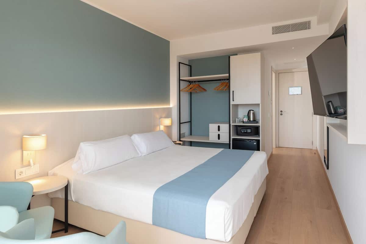 Quarto de hotel moderno com cama, mesas de cabeceira, escrivaninha, cadeira, armário e cozinha compacta.