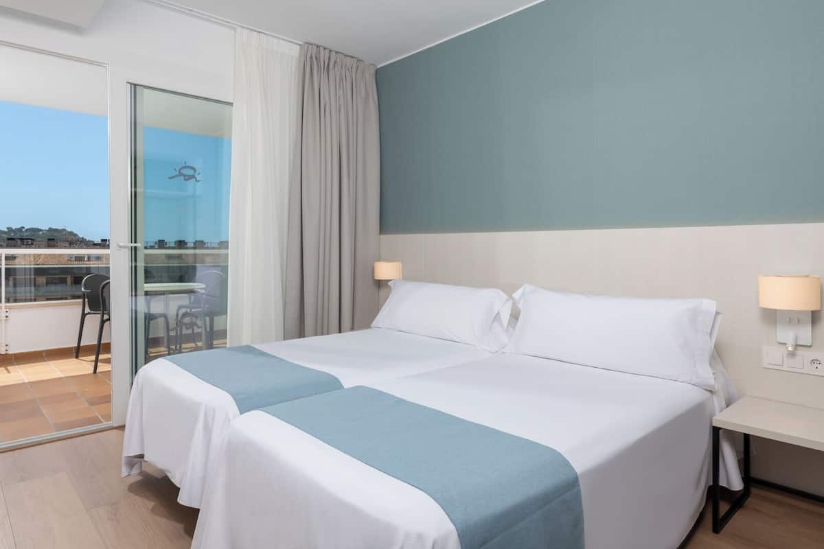 Quarto de hotel com cama, duas mesas de cabeceira, uma cadeira, porta de vidro que dá para uma varanda e vista para a cidade.