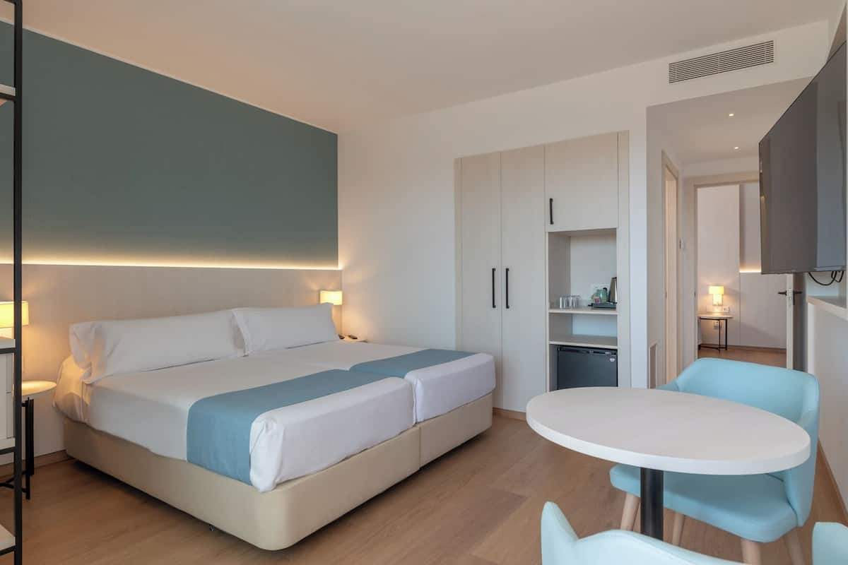 Quarto de hotel moderno com cama, escrivaninha, cadeiras e televisão.