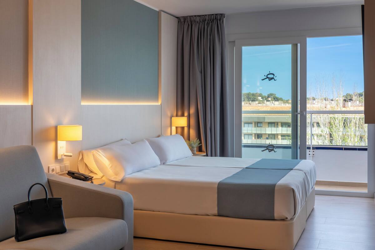 Quarto de hotel com cama, mesa de cabeceira, luminária, poltrona e vista para a cidade pela janela.
