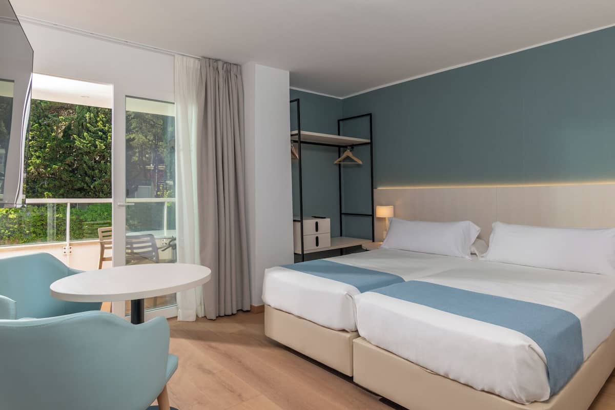 Quarto de hotel com uma cama grande, uma mesinha, uma cadeira, varanda com vista para o verde e um guarda-roupa.