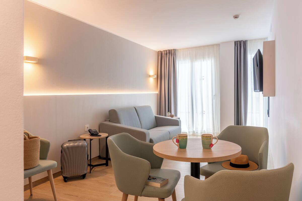 Quarto de hotel moderno com sofá, mesa de jantar com cadeiras, uma mesinha lateral, mala e televisão fixada na parede.