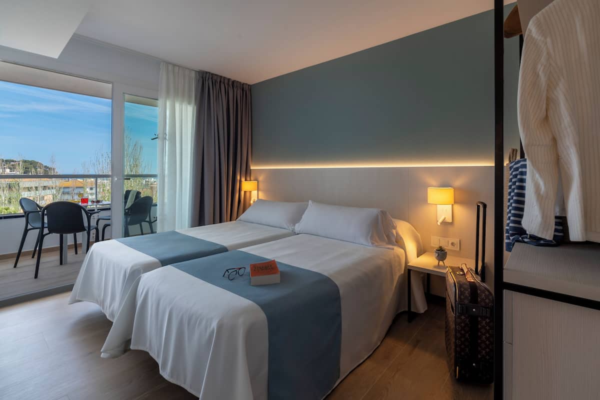 Quarto de hotel com duas camas, uma escrivaninha e uma cadeira. Janelas grandes oferecem vista para a cidade.