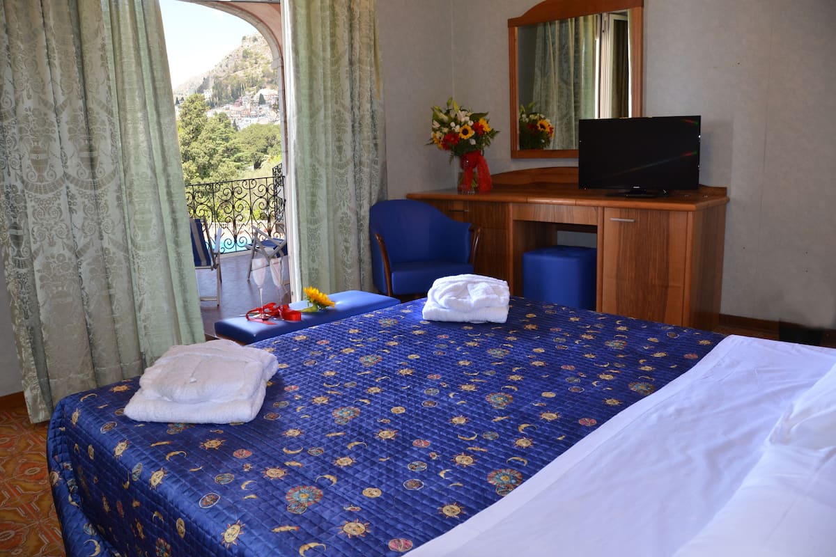 Quarto de hotel com cama, escrivaninha com televisão, cadeira e vista para uma paisagem montanhosa.