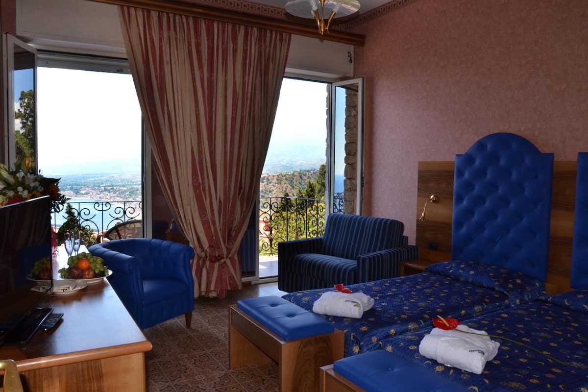 Quarto de hotel com cama, escrivaninha, cadeira, varanda com vista e mesa de jantar com frutas.