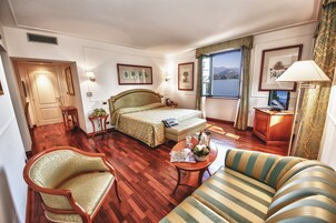 Quarto de hotel com cama, sofá, cadeira, mesa com abajur e janela com vista.