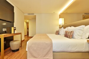 Quarto de hotel com cama, mesa de cabeceira, abajur e um banquinho.