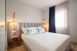 Uma cama bem arrumada com almofadas listradas, uma mesa de cabeceira com abajur e uma janela com cortinas.