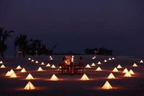 Um casal jantando à mesa, cercado por diversas luzes cônicas numa praia à noite.
