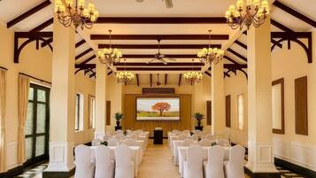Um salão de banquetes com fileiras de mesas e cadeiras cobertas de branco, lustres e uma tela grande na frente.
