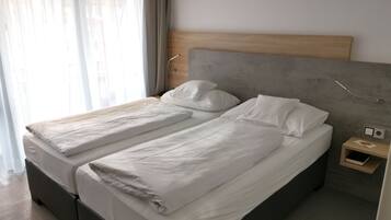 Uma cama de casal com cabeceira de madeira, uma mesa de cabeceira com abajur e uma janela com cortinas leves.