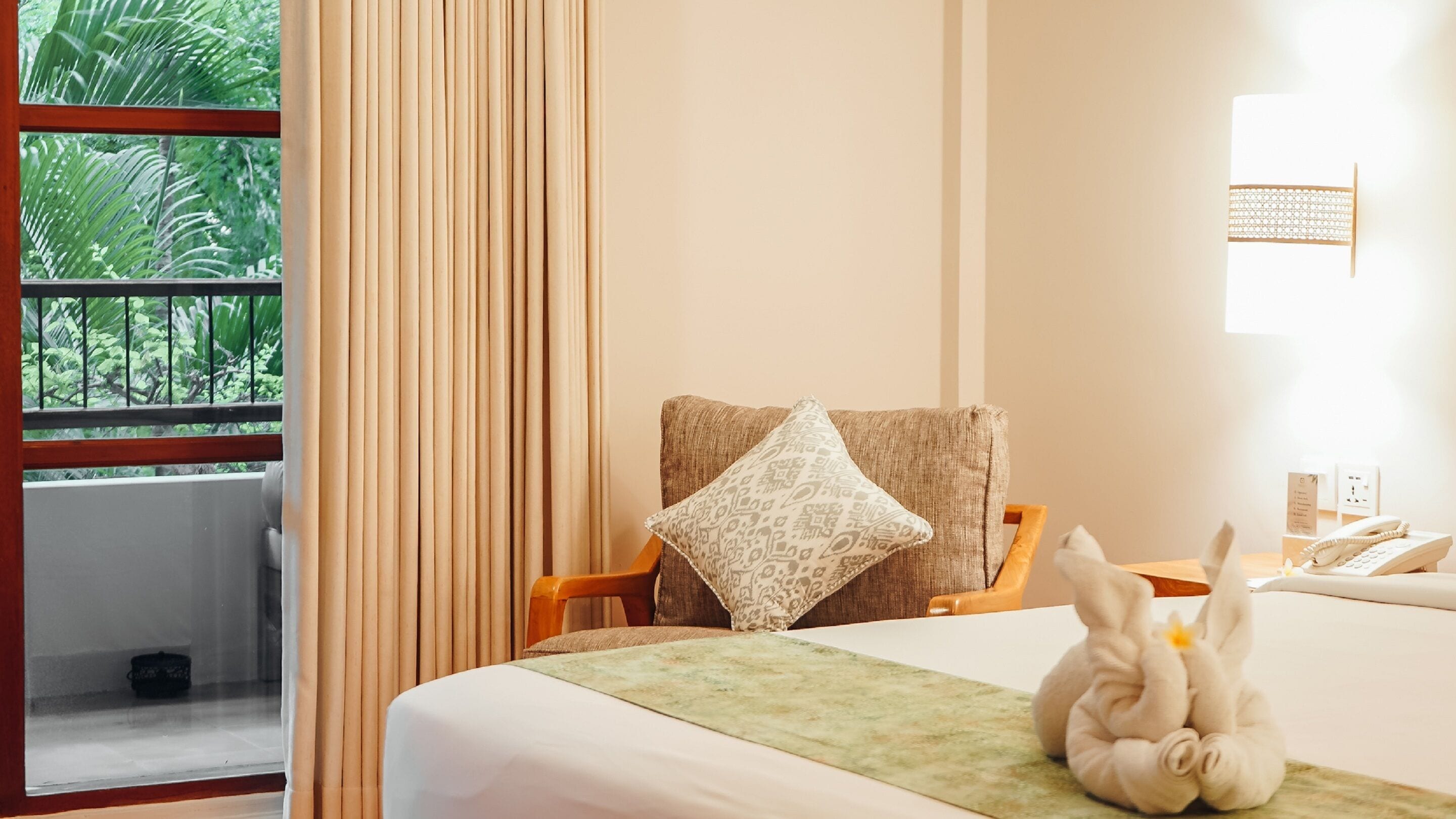 Quarto de hotel com cama, uma cadeira, janela com cortinas, telefone fixado na parede e vista para o verde lá fora.