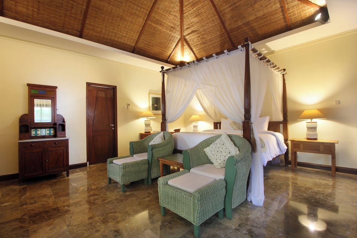 Quarto com cama de dossel, duas cadeiras de vime verde, mesa de cabeceira de madeira com abajur e um armário de madeira.