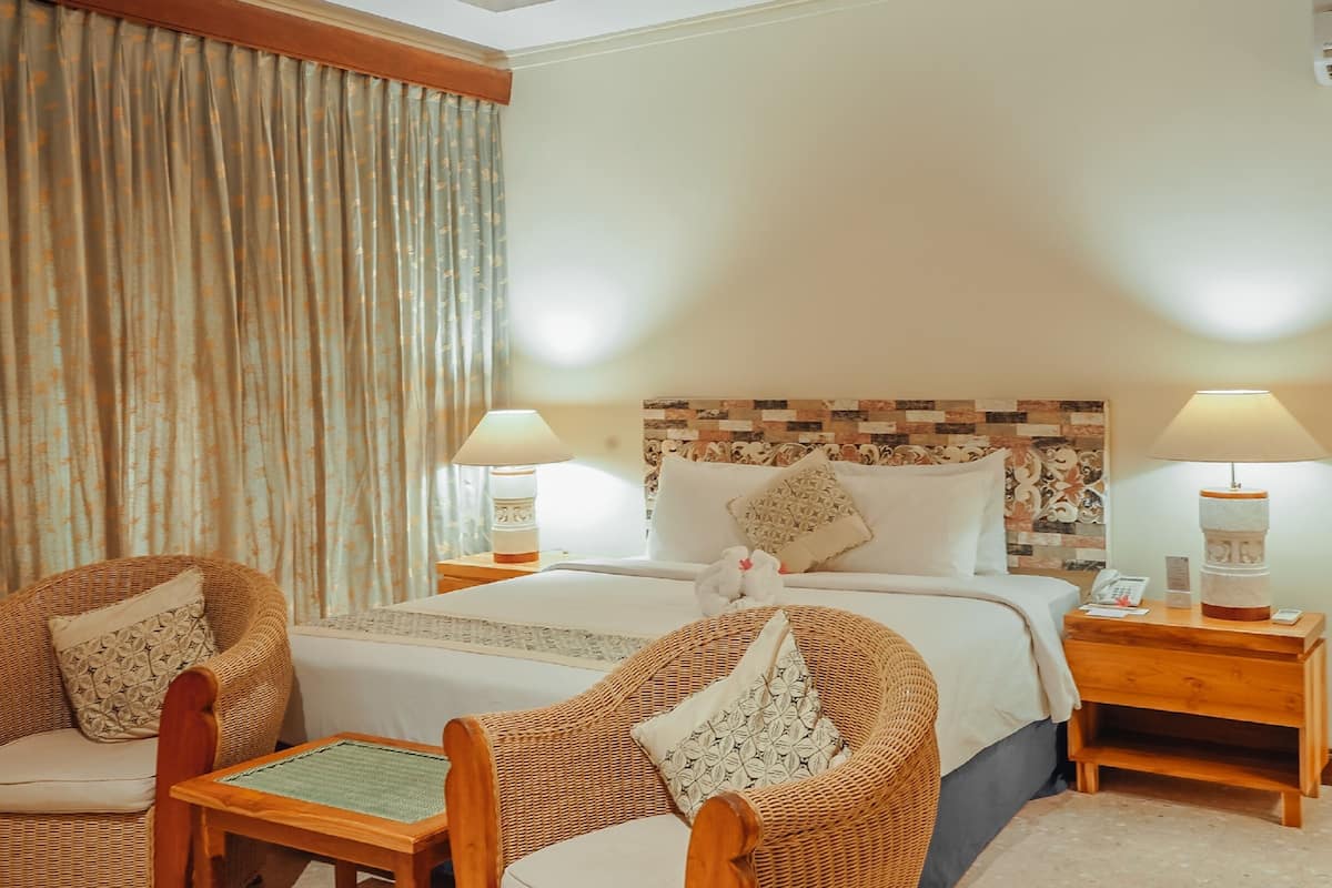 Quarto de hotel com uma cama, duas cadeiras de vime, uma mesa pequena e duas luminárias de cabeceira.