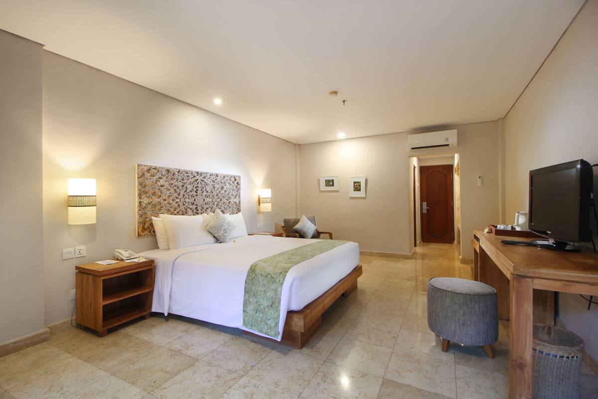 Quarto de hotel com uma cama grande, mesas de cabeceira, uma escrivaninha com televisão e um banquinho.