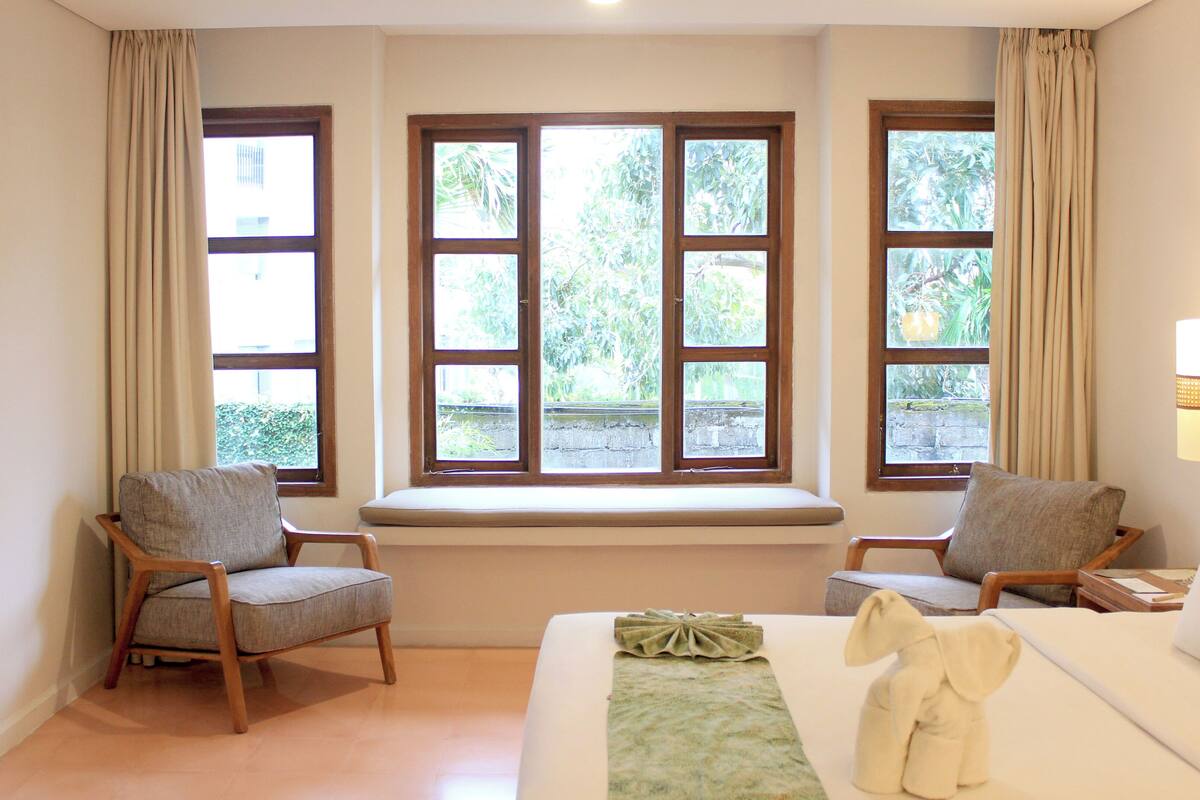 Quarto de hotel com uma cama grande, duas poltronas, um banco e três janelas com vista para o verde.
