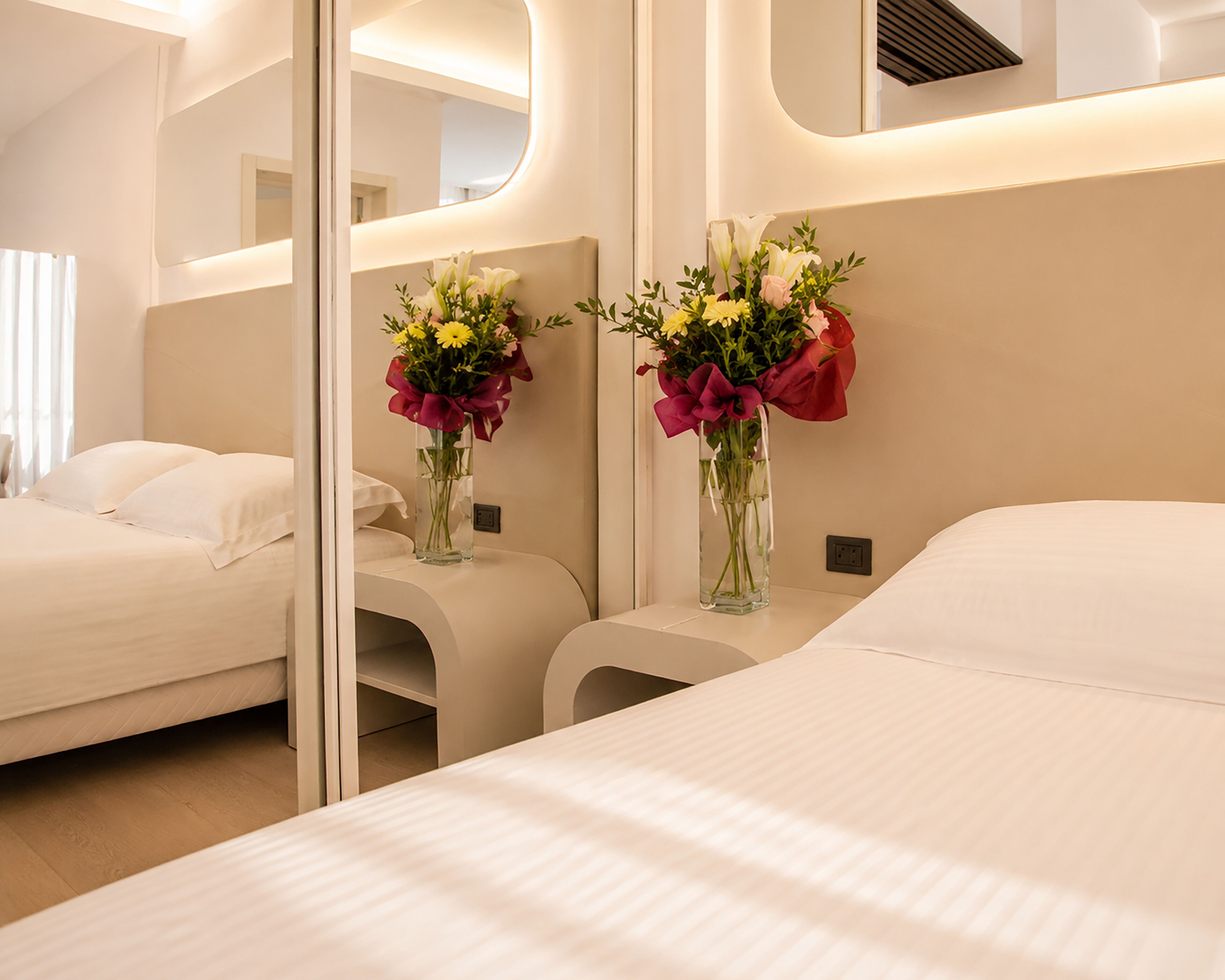 Quarto de hotel com uma cama grande, duas mesas de cabeceira e um espelho. Há dois vasos com flores sobre as mesas de cabeceira.