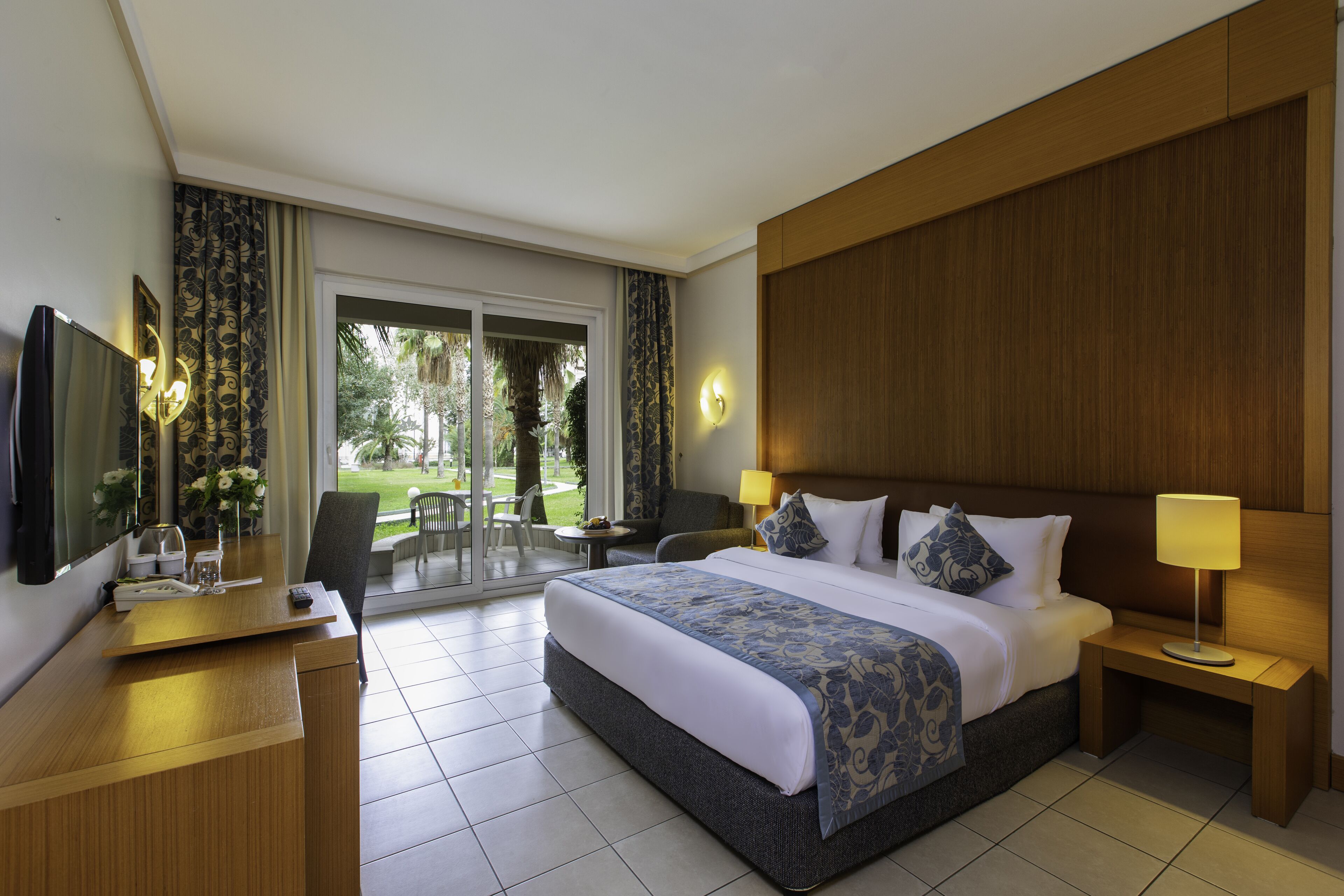 Quarto de hotel com uma cama grande, uma escrivaninha com computador, uma televisão e vista para o exterior.