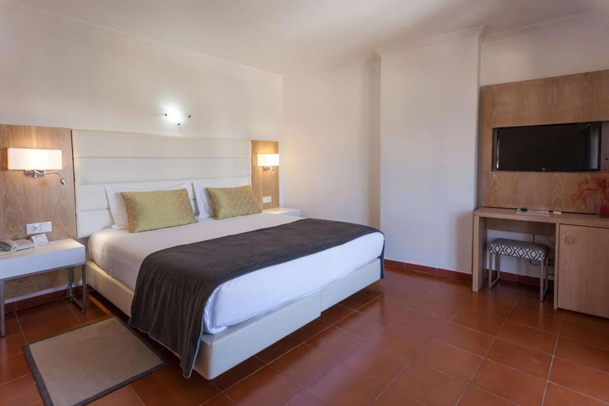 Quarto de hotel com cama, mesas de cabeceira, escrivaninha, cadeira, televisão e luminária fixa na parede.