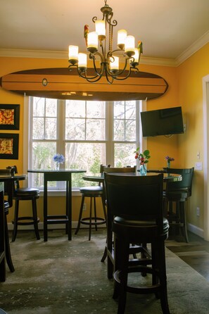 Uma sala de jantar com um candelabro, uma televisão fixada na parede e uma janela com vista para árvores.