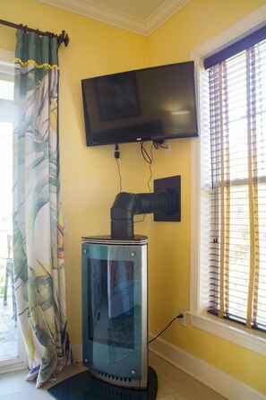 Quarto com televisão fixada na parede, aquecedor no chão, janela com persianas e cortina estampada com folhas.