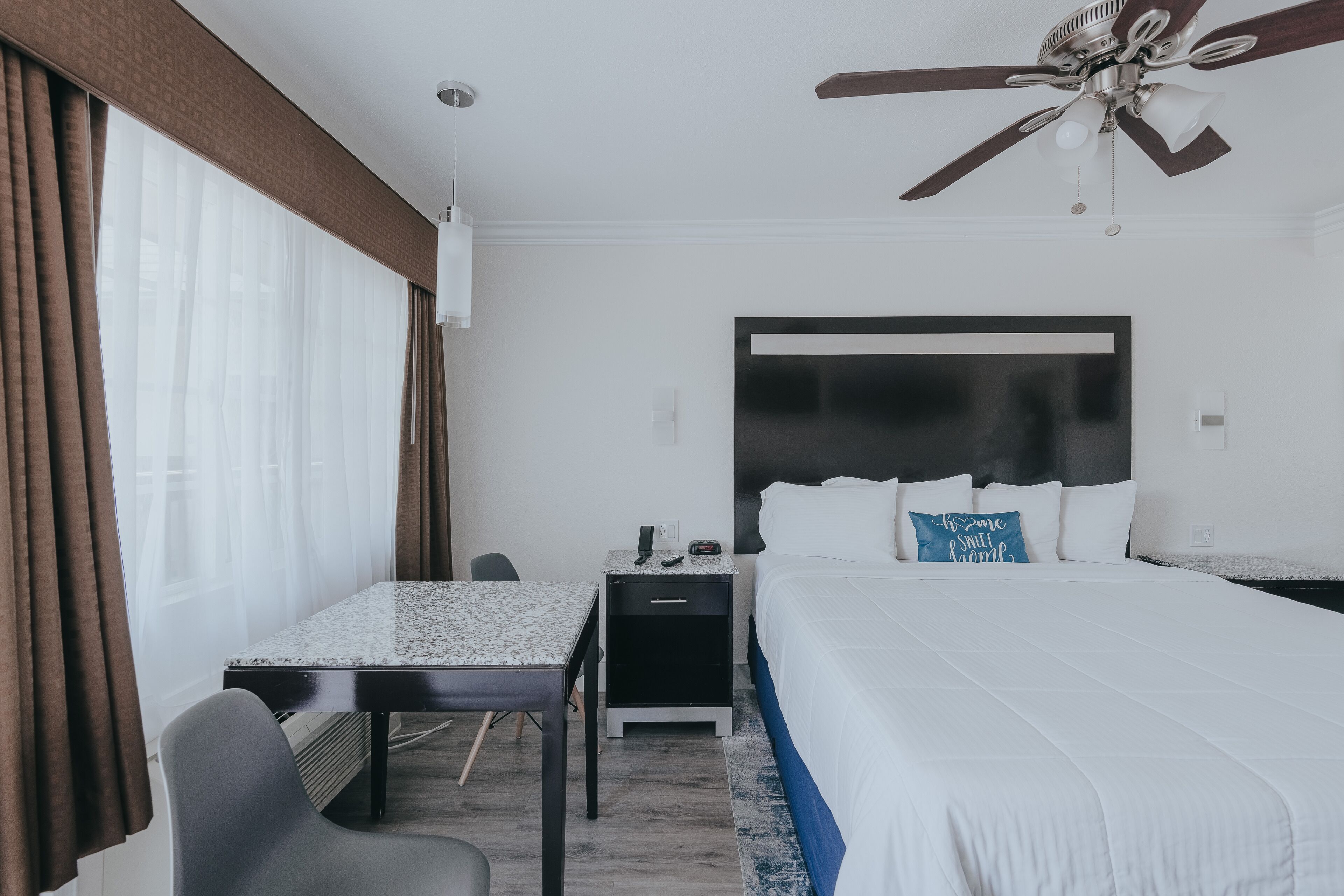 Quarto de hotel com cama, escrivaninha, cadeira, mesa de cabeceira, ventilador de teto e janela com cortinas.