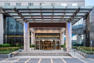 Entrada de hotel moderna com toldo de vidro, porta giratória e área externa bem cuidada, com degraus e jardineiras.