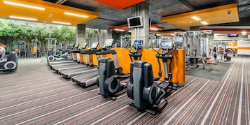 Uma academia com várias esteiras, aparelhos de musculação e uma tela de televisão.