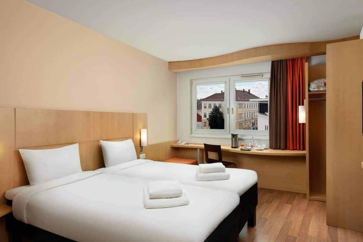 Quarto de hotel com duas camas, uma escrivaninha, uma cadeira e uma janela com cortinas.