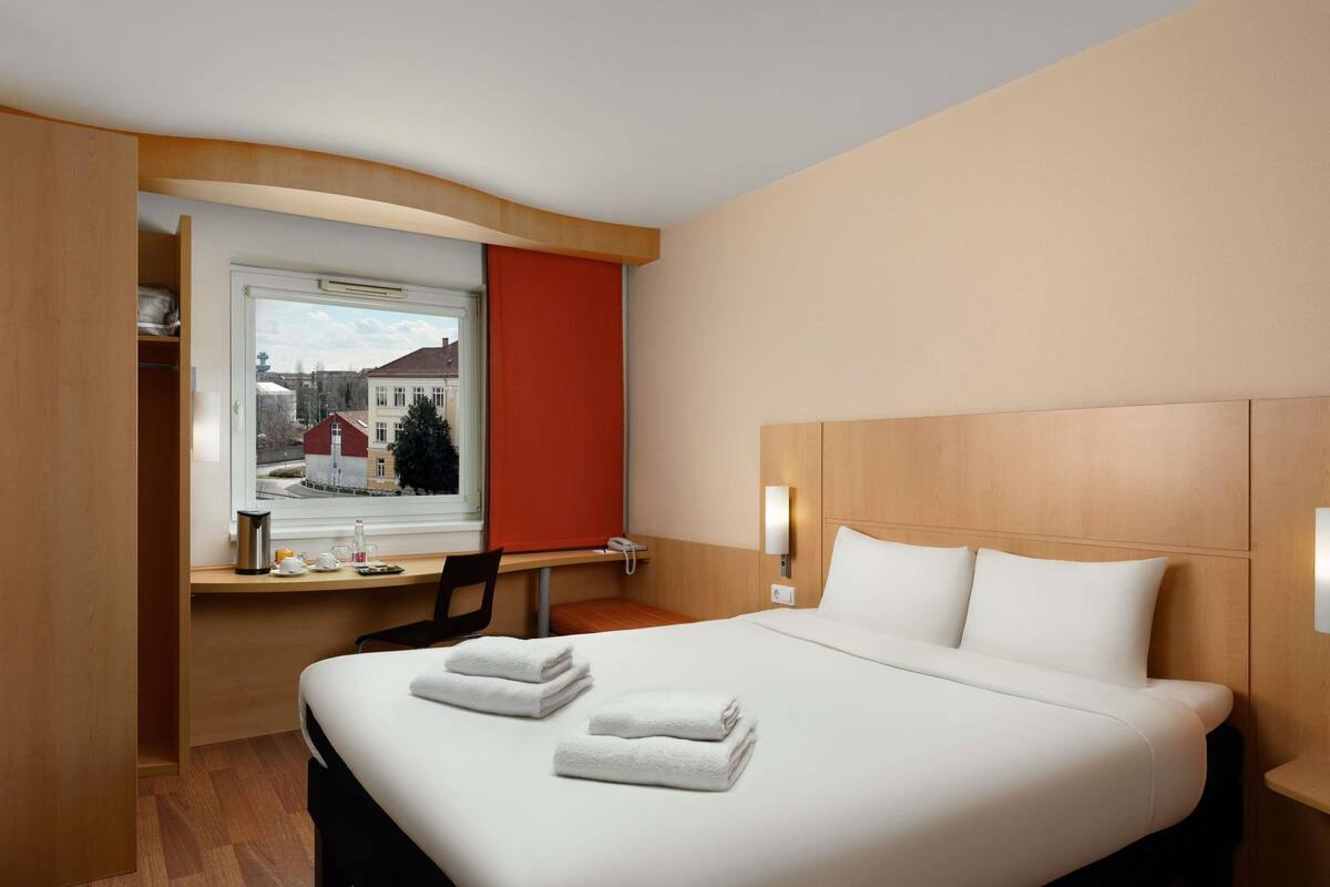 Quarto de hotel com uma cama grande, uma escrivaninha com cadeira, uma janela com vista para edifícios e uma cortina vermelha.