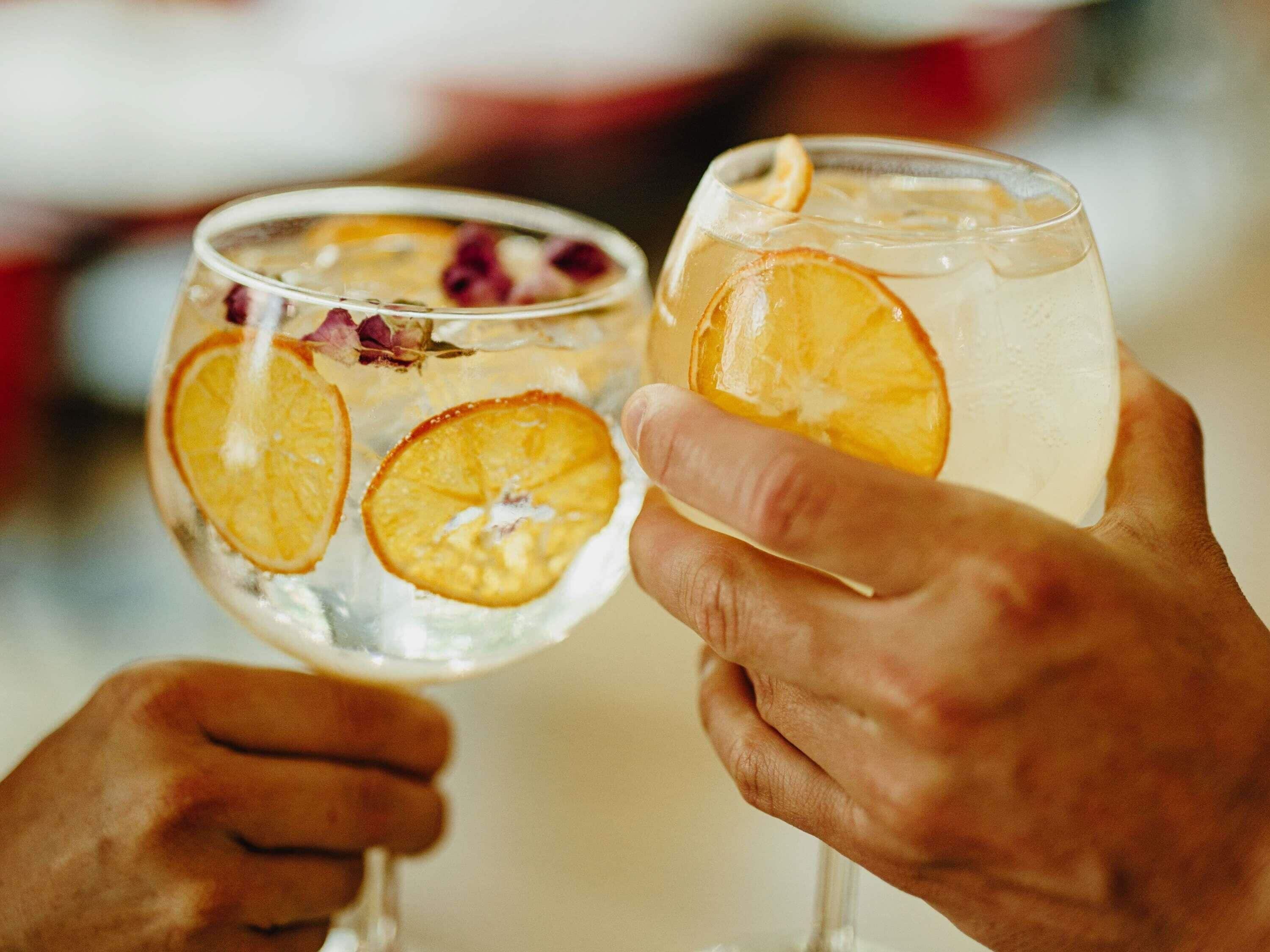 Duas mãos segurando copos com bebida e fatias de laranja.