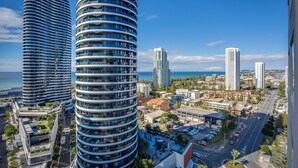Um edifício alto com fachada curva, cercado por outros edifícios altos, um estacionamento e vista para o mar.