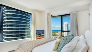 Quarto de hotel moderno com uma cama grande, uma televisão num criado-mudo e vista para a cidade através de amplas janelas.
