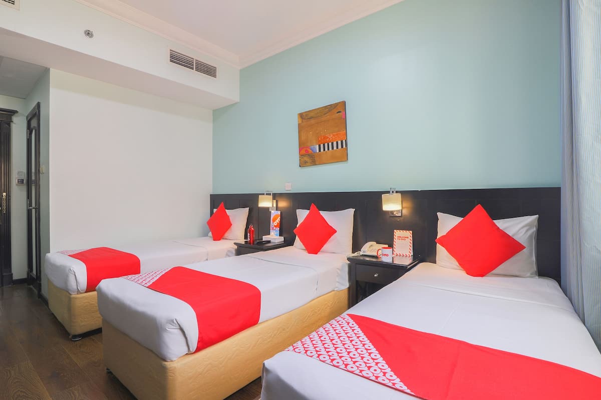 Quarto de hotel com três camas, cada uma com roupa de cama vermelha e branca, uma mesa de cabeceira com telefone e uma obra de arte na parede.
