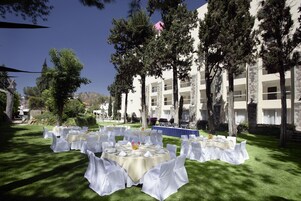 Um espaço para eventos ao ar livre com mesas redondas dispostas para refeições, rodeado por cadeiras brancas, em uma área de jardim bem cuidada com árvores e um edifício ao fundo.
