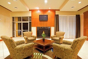 Um hall de hotel com lareira, uma TV de tela plana e quatro poltronas estampadas ao redor de uma mesa de centro de madeira.