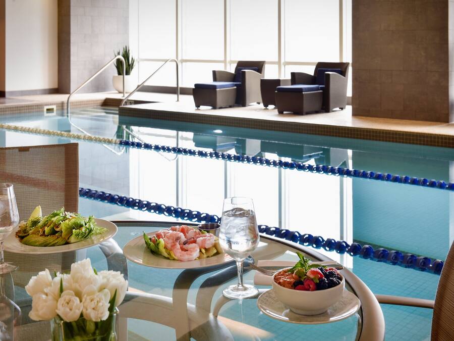 Uma área de refeições à beira da piscina, com uma mesa posta para dois, apresentando uma salada, camarões e uma cesta de frutas.