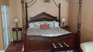 Uma cama de dossel com estrutura de madeira e cabeceira com design curvo. Há duas mesas de cabeceira com abajures de cada lado da cama.