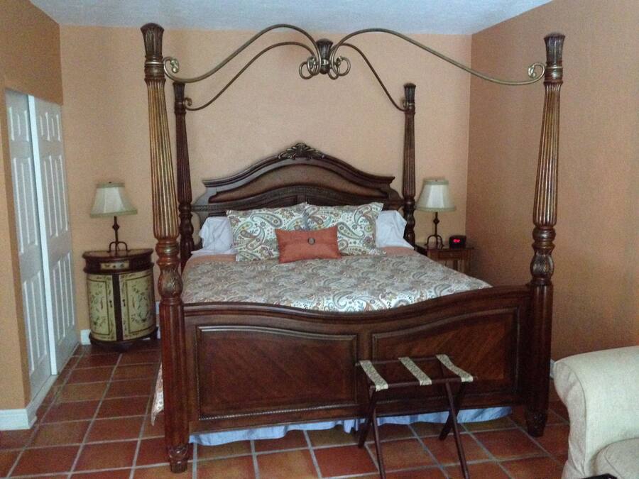 Uma cama de dossel com estrutura de madeira e cabeceira com design curvo. Há duas mesas de cabeceira com abajures de cada lado da cama.